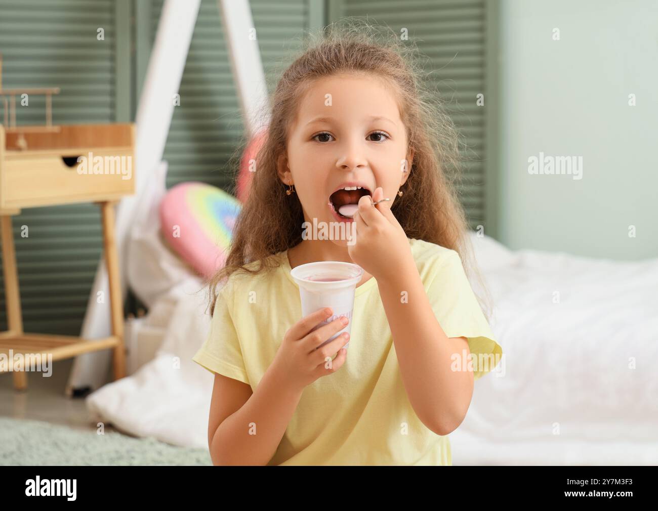 Cute little girl with spoon eating yogurt in room Stock Photo - Alamy