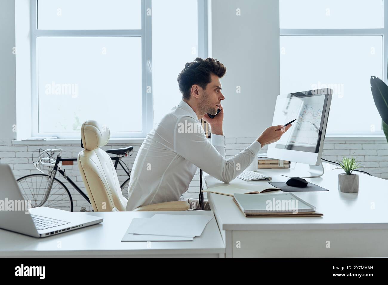 Confident young man pointing computer monitor and talking on phone ...