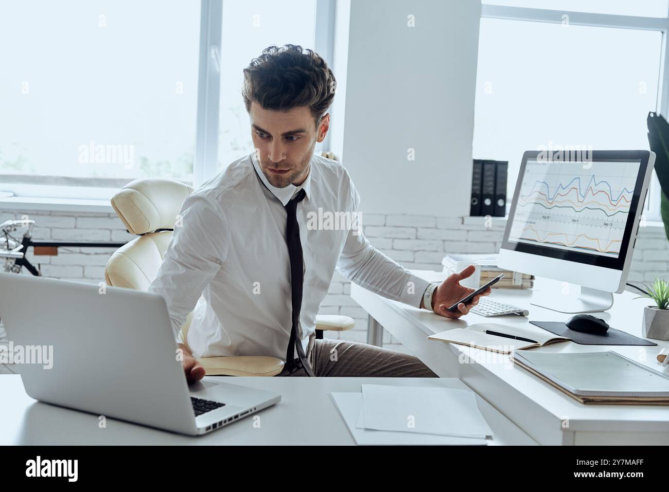 Confident young man using computer and holding smart phone while ...