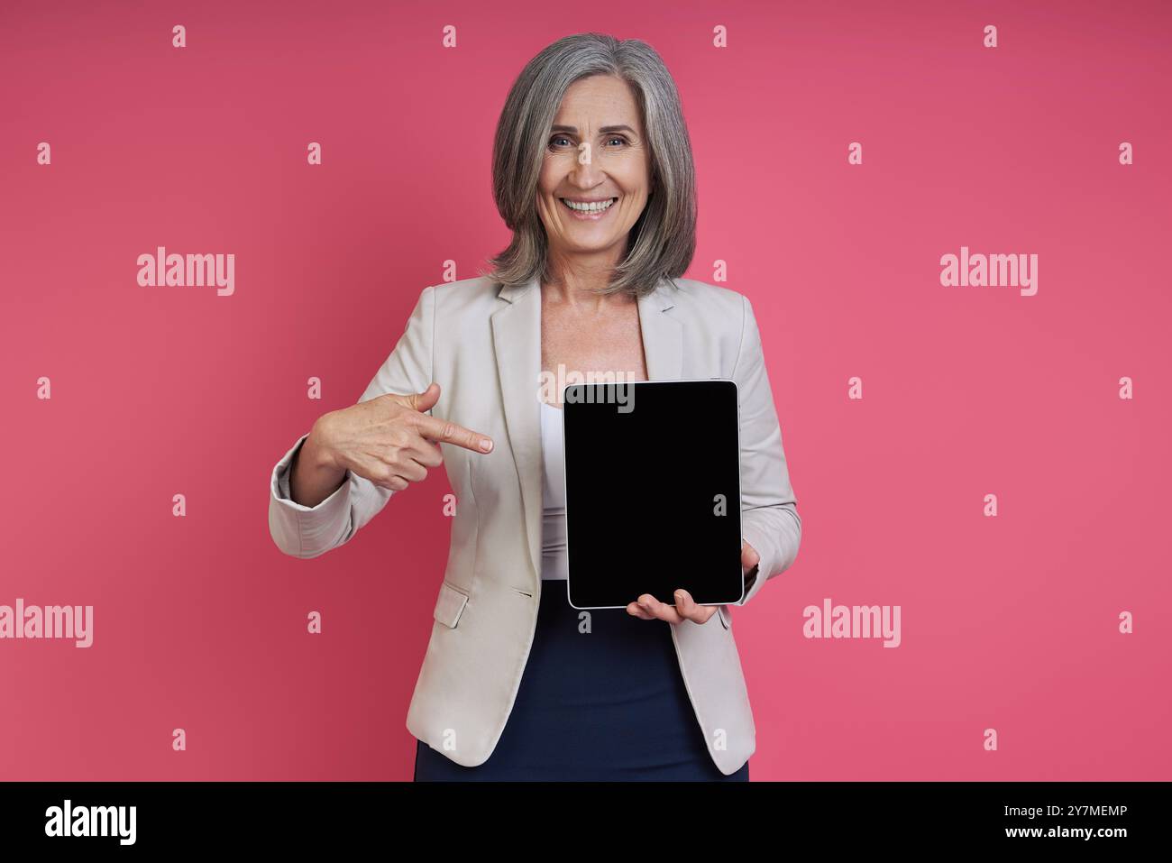 Happy senior woman in formalwear pointing digital tablet while standing ...