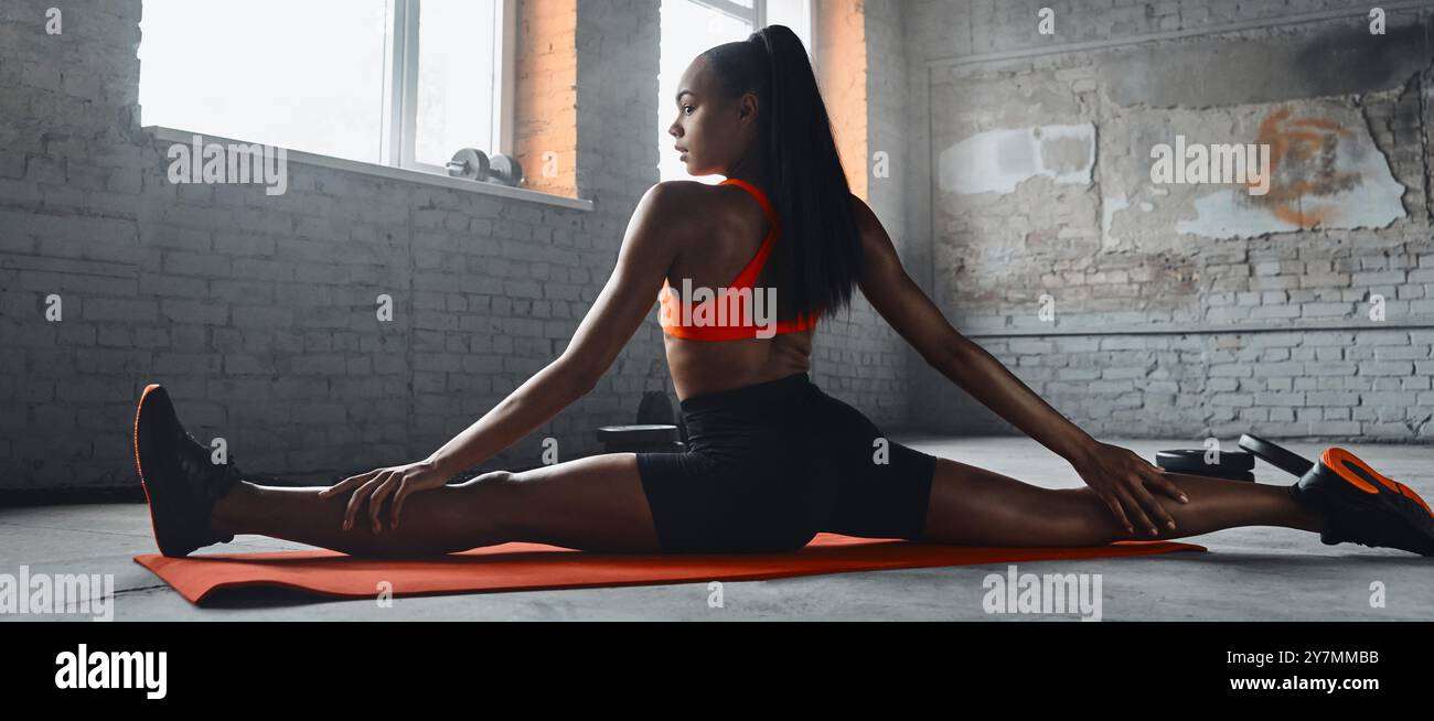 Rear view of attractive young African woman stretching on splits in gym ...