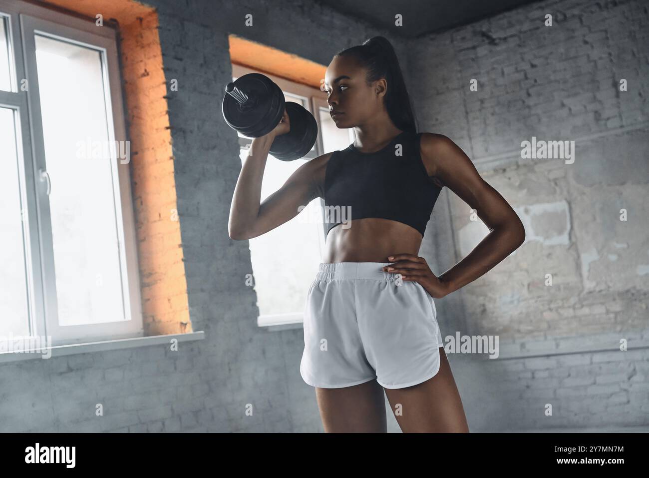 Concentrated young African woman using dumbbell while exercising in gym ...