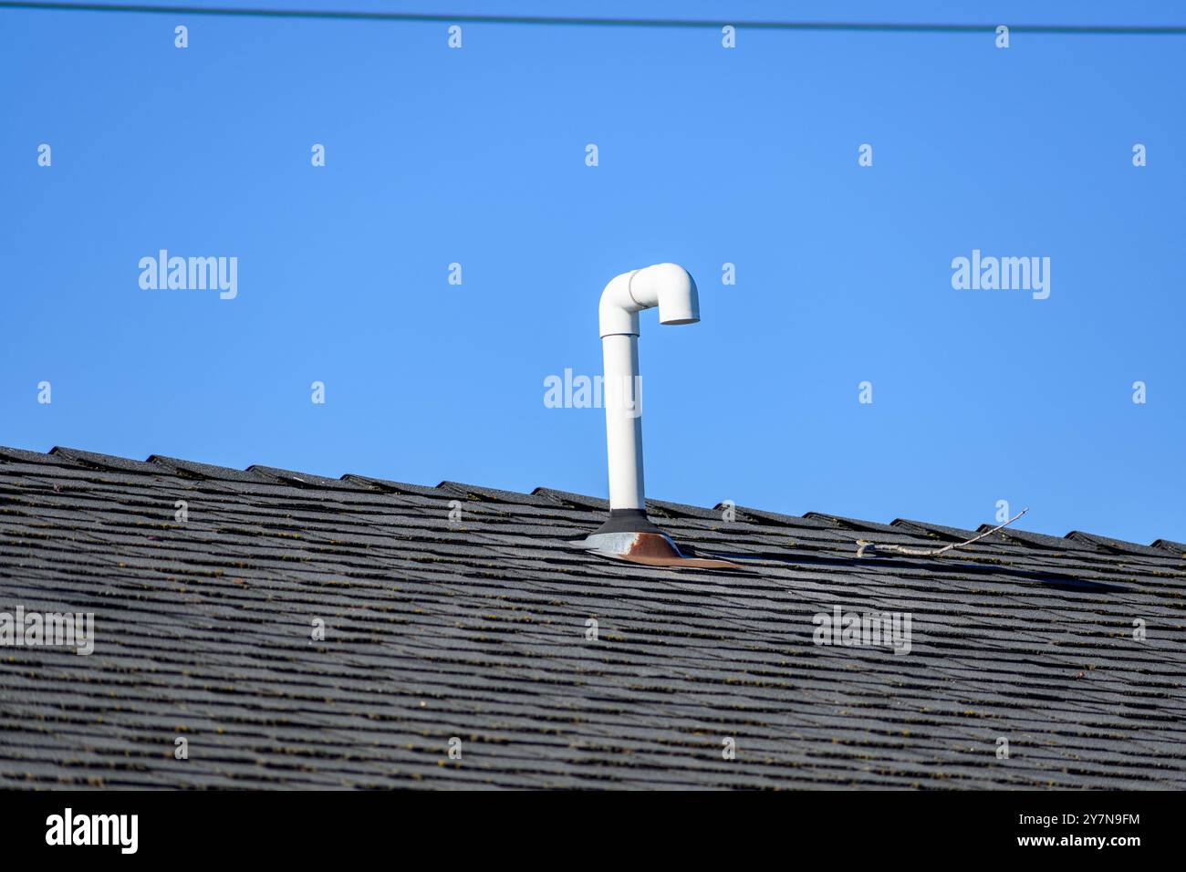 A white PVC pipe vent protrudes from a dark shingle roof, set against a ...