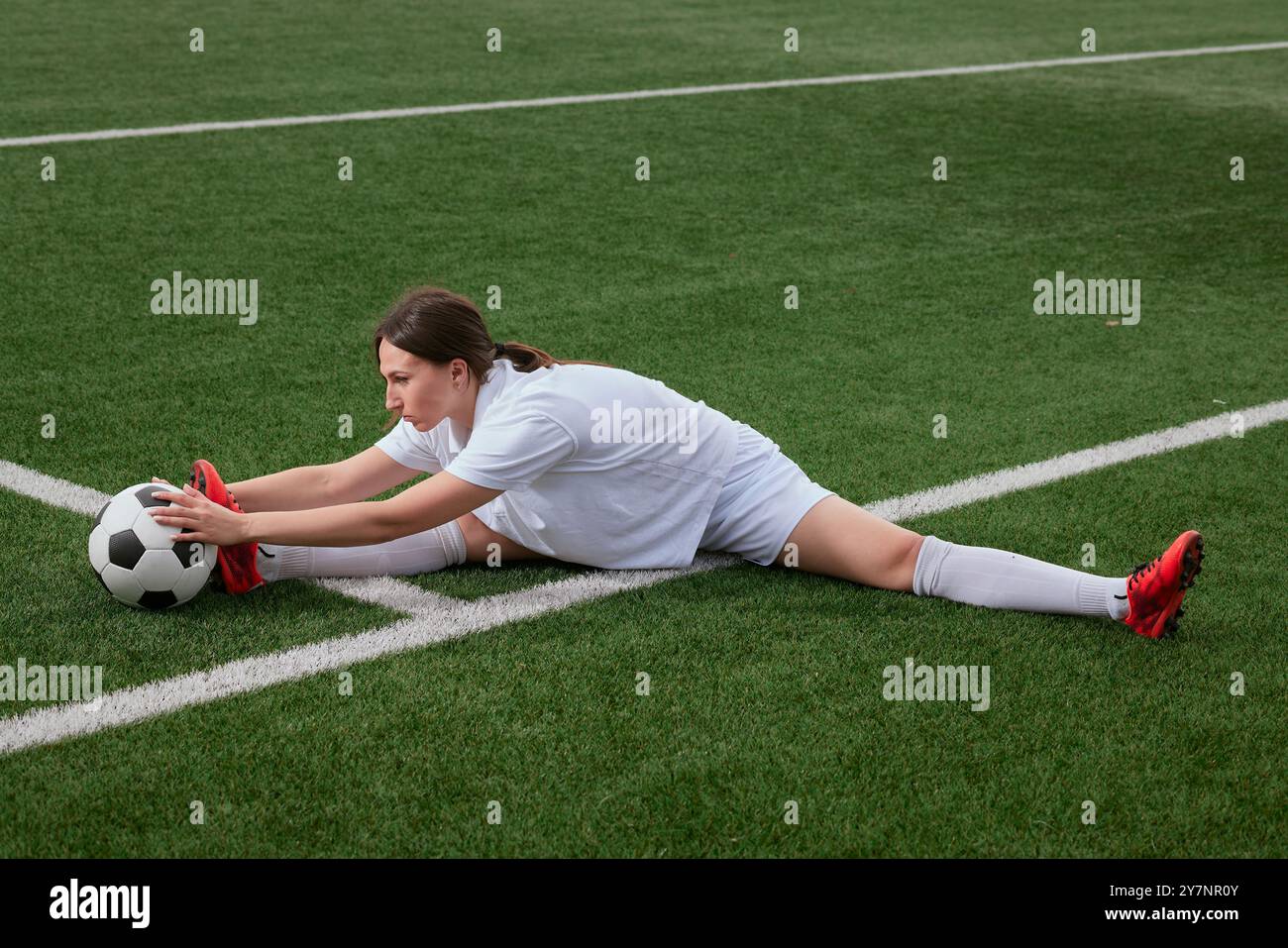 A young soccer player stretches on a green field. She is in a split ...