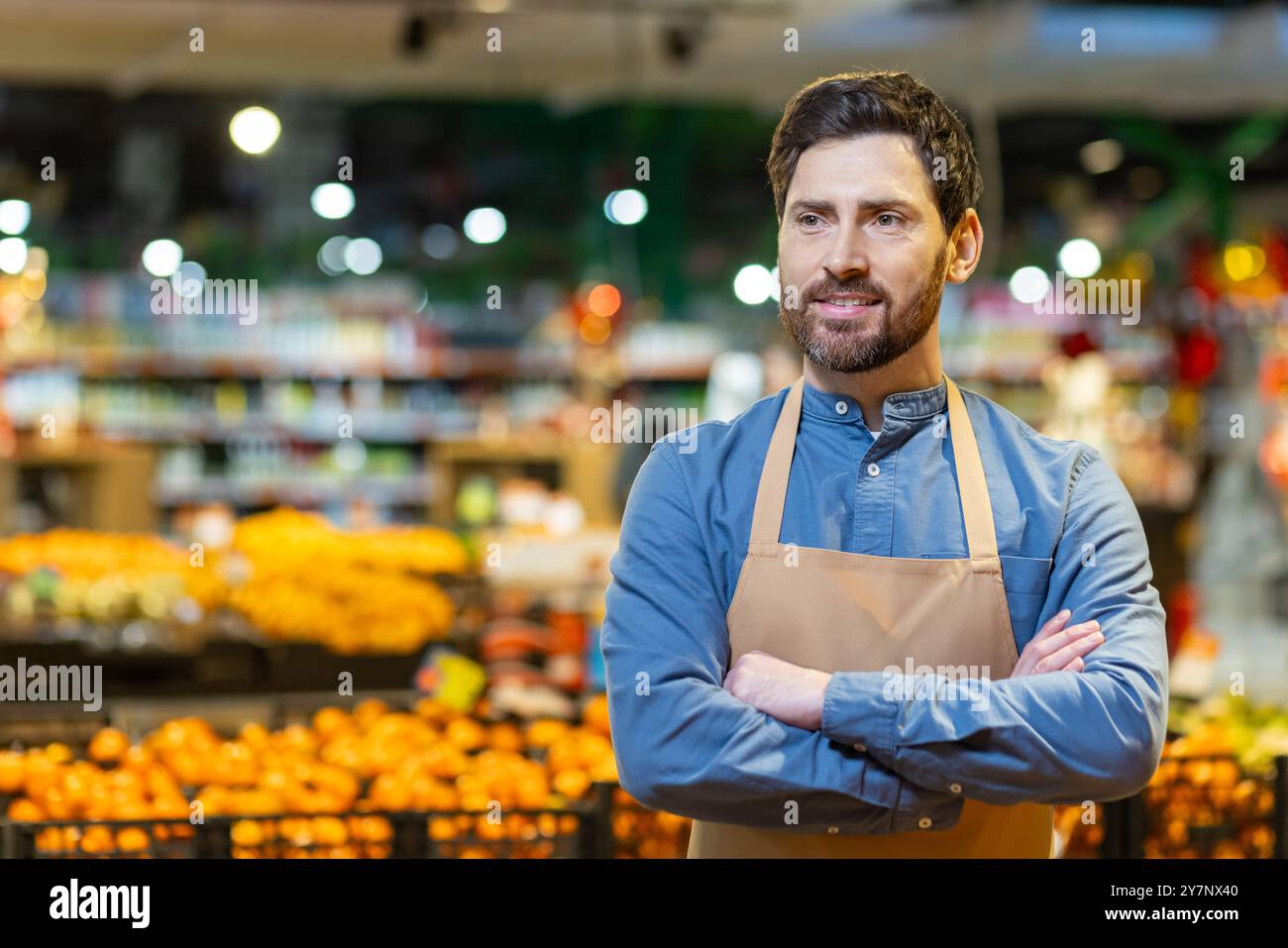 Grocery store owner in apron stands confidently with arms crossed in ...