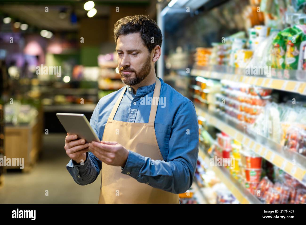 Supermarket employee wearing apron uses tablet in grocery aisle. Man ...