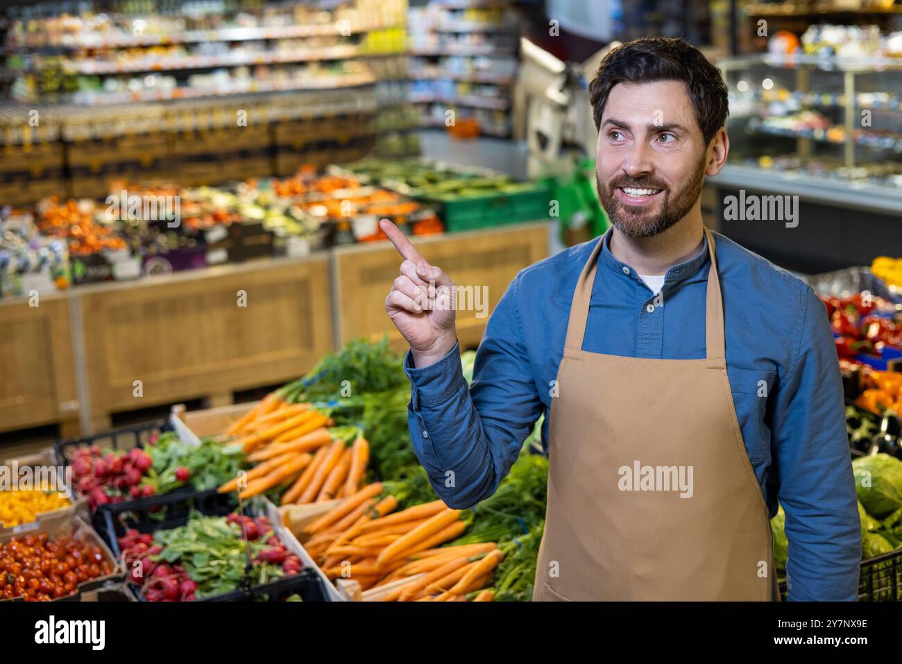 Grocery store worker wearing apron smiles while pointing towards fresh ...