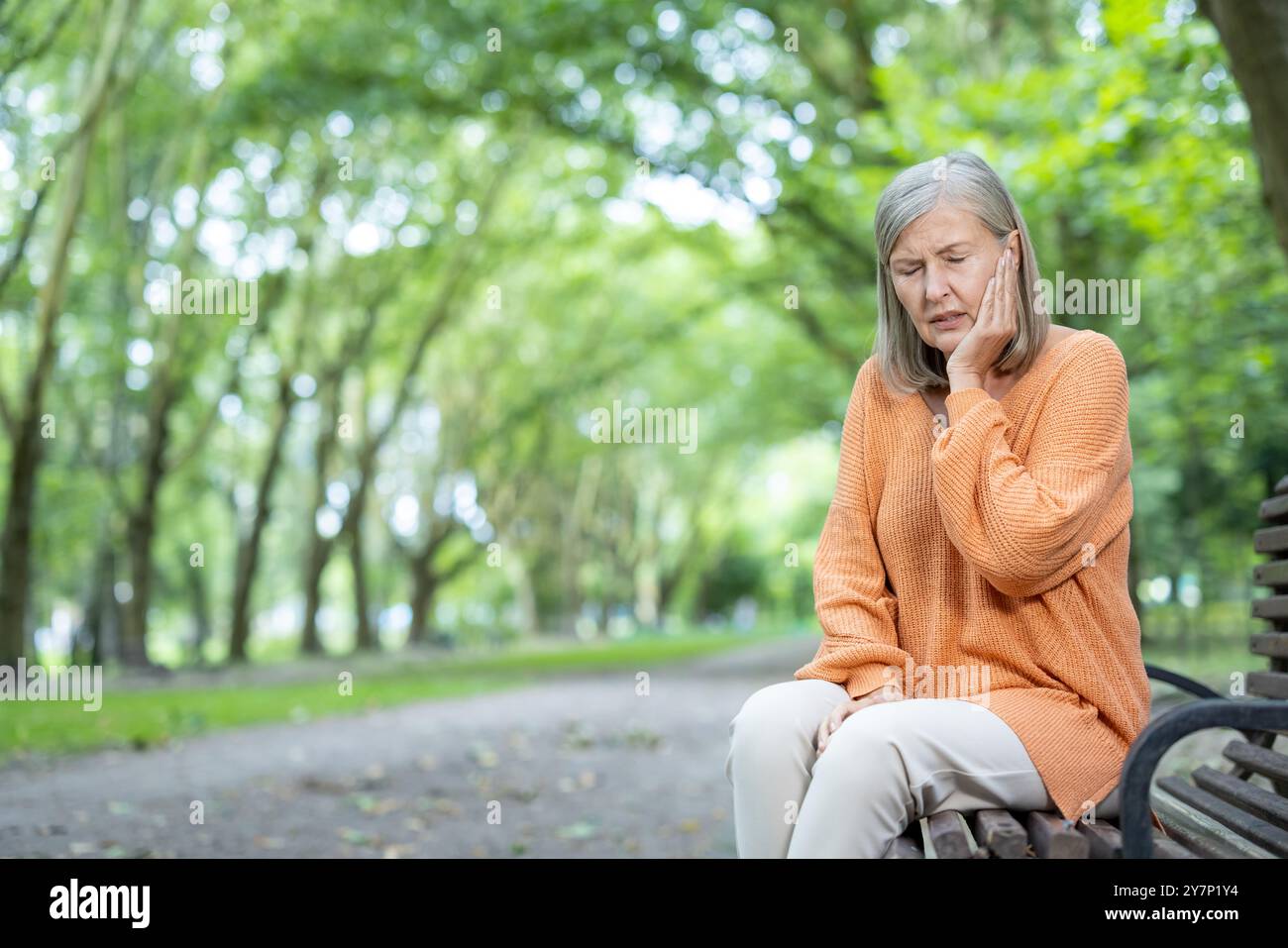 Senior woman suffering from toothache, sitting outdoors on a bench with ...
