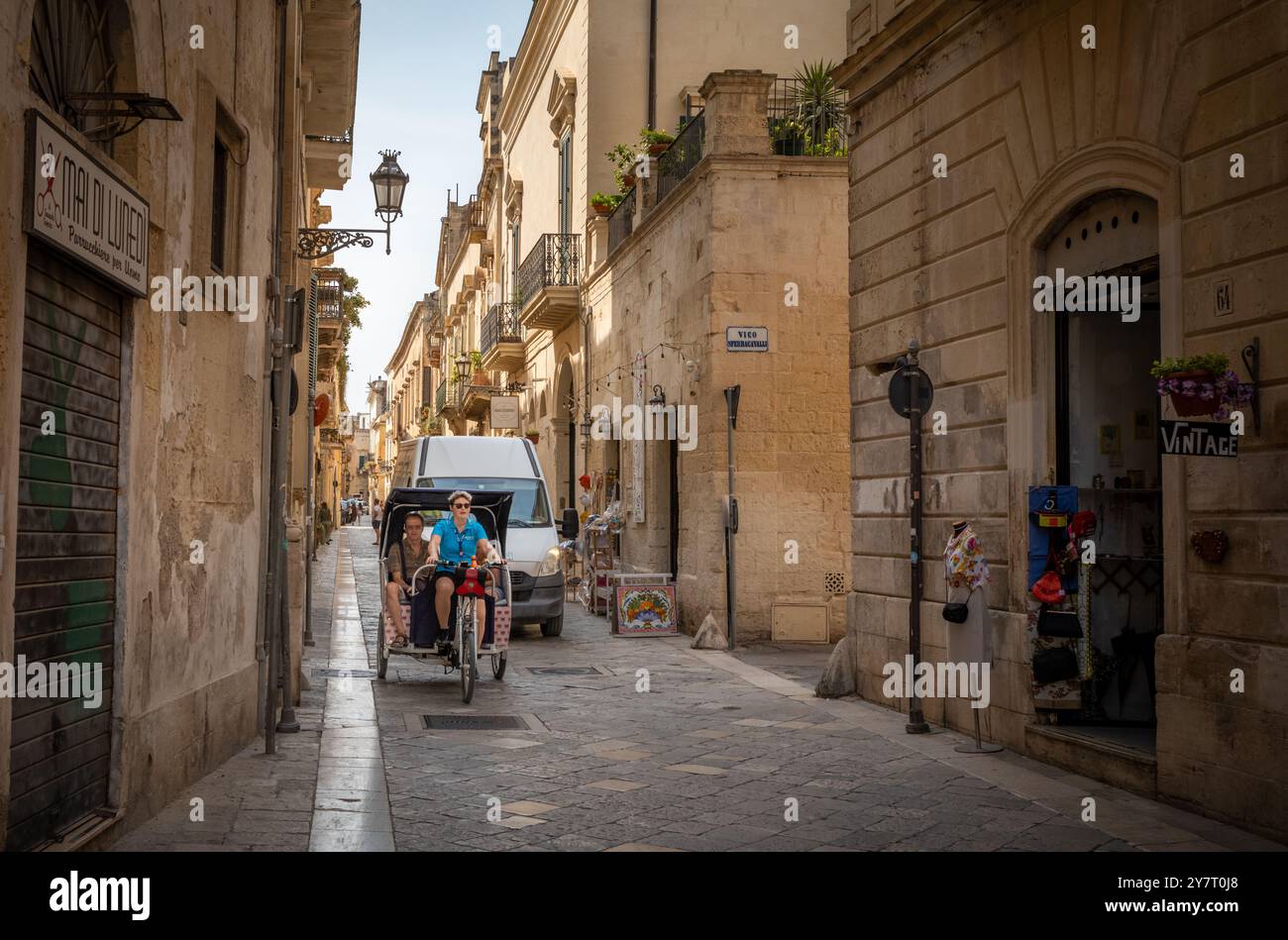 A rickshaw pedicab is followed by a white van down a narrow street in ...