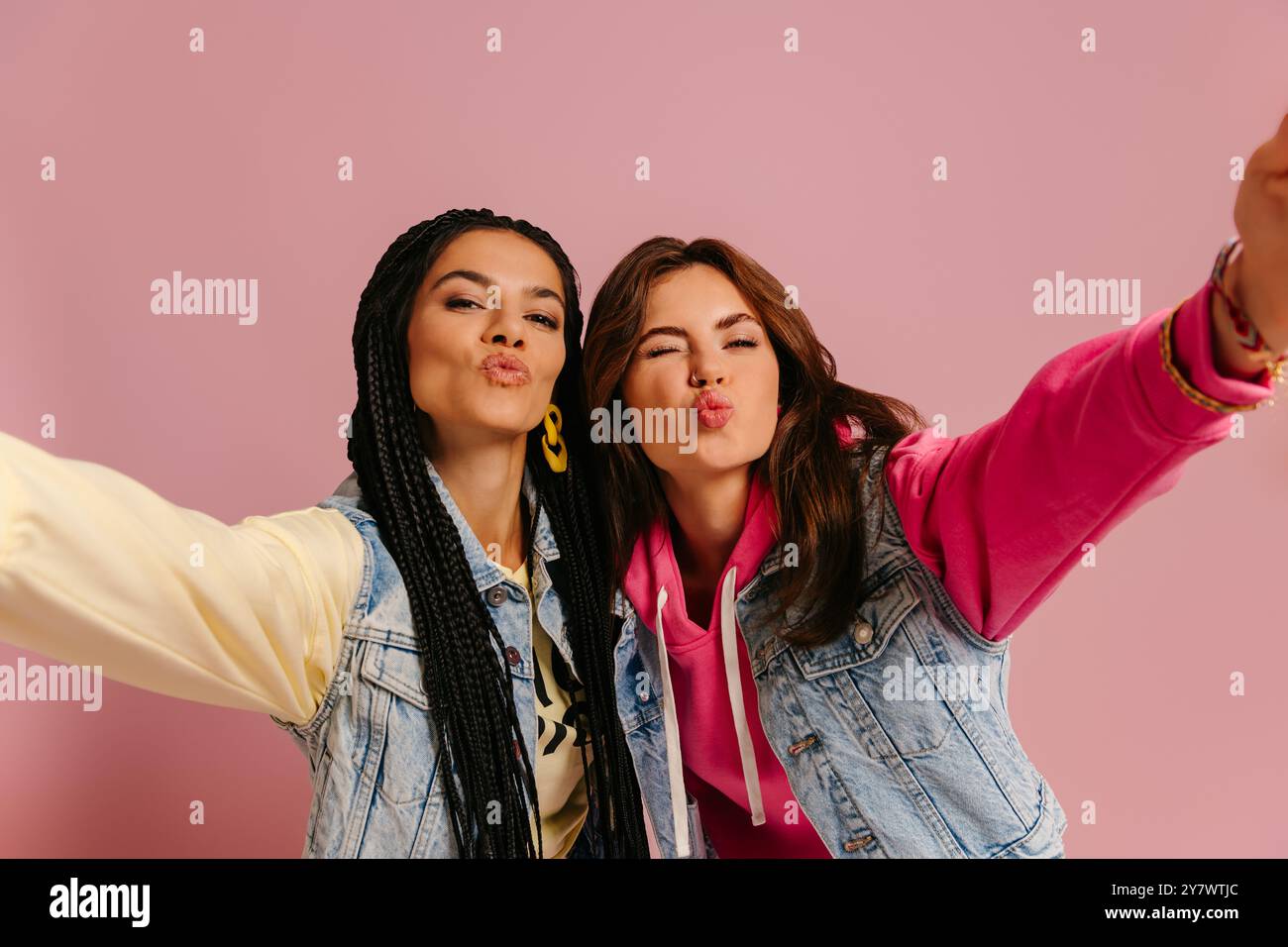 Two playful young women in colorful wear embracing and making self portrait against pink ...