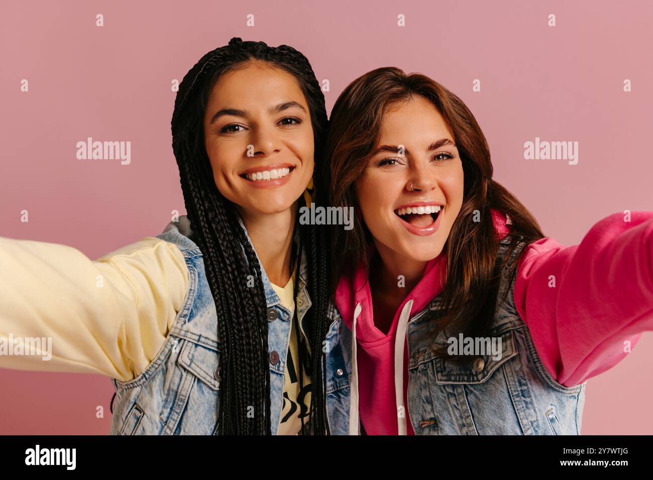 Two joyful young women in colorful wear embracing and making self portrait against pink ...
