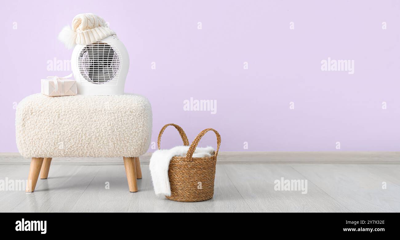 Electric fan heater, hat, gift box, pouf and basket near lilac wall ...