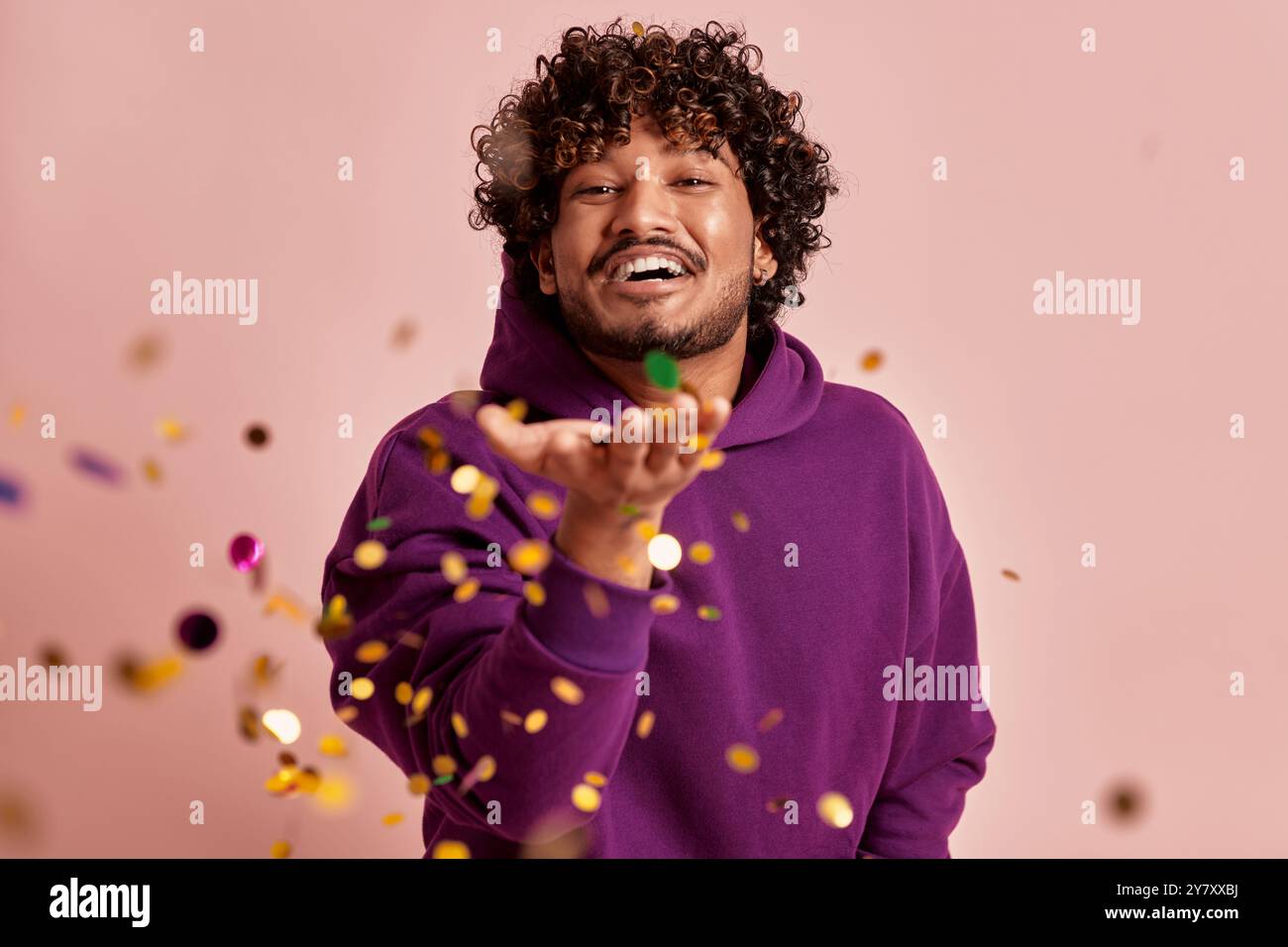 Happy Indian man blowing colorful confetti while standing against pink ...