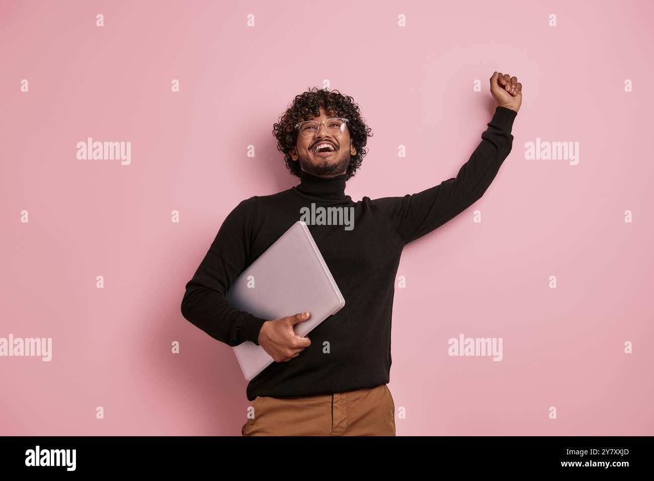 Joyful Indian man carrying laptop and gesturing against pink background ...