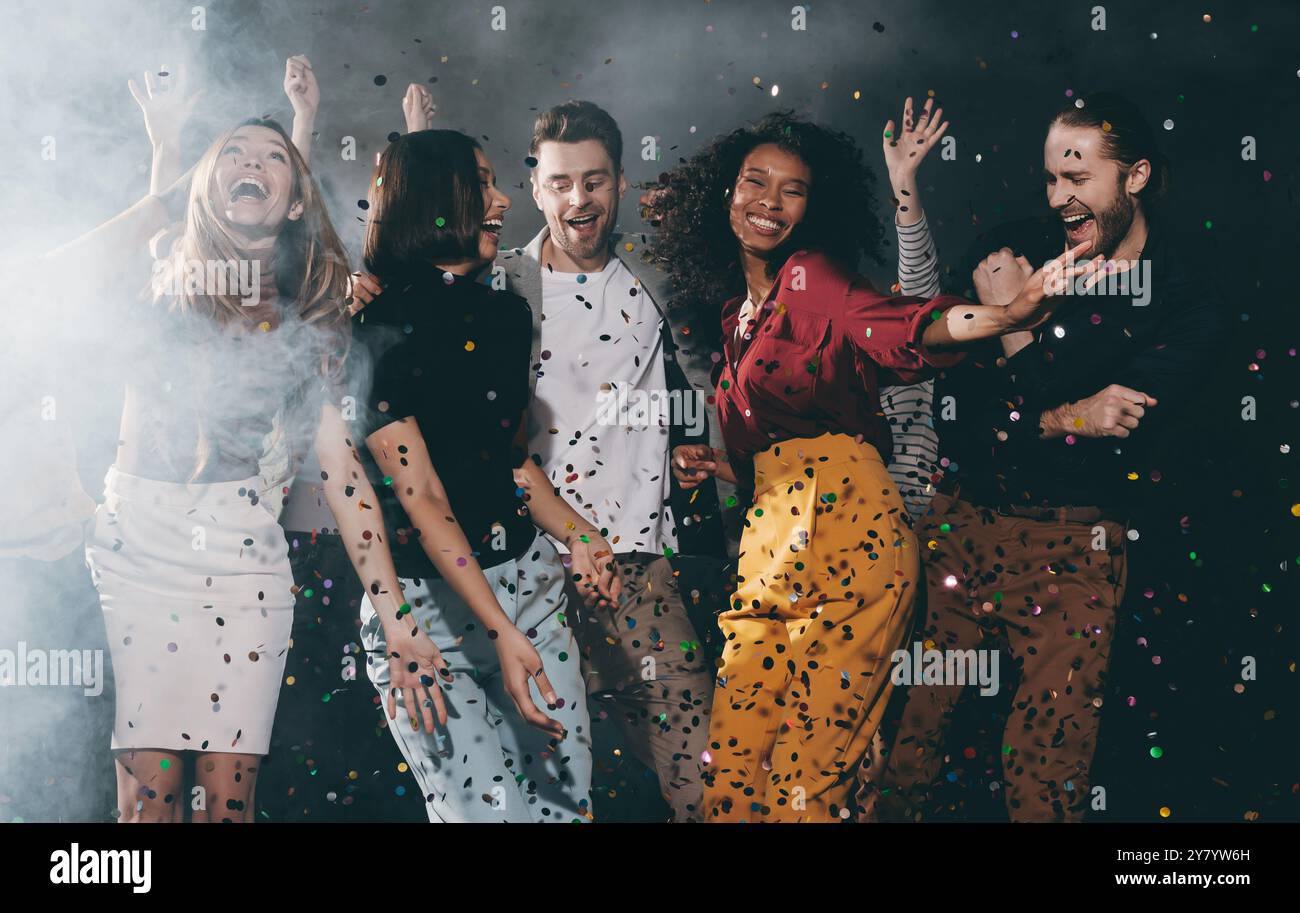 Group of joyful young friends dancing and throwing confetti while ...