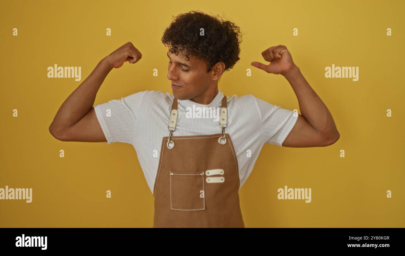 Young man flexing muscles in a brown apron over an isolated yellow ...
