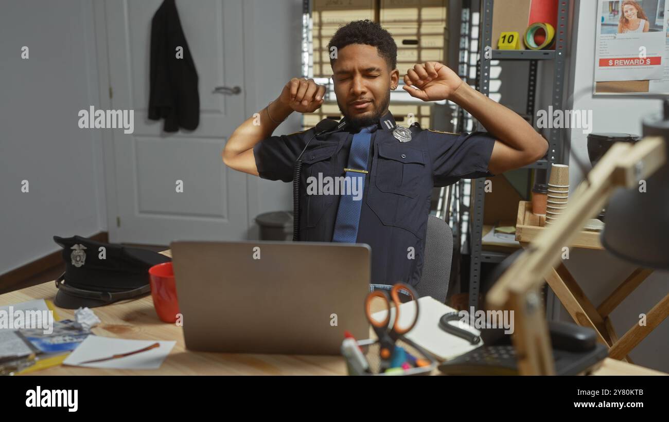 Tired black policeman stretching at office desk, with laptop, uniform ...