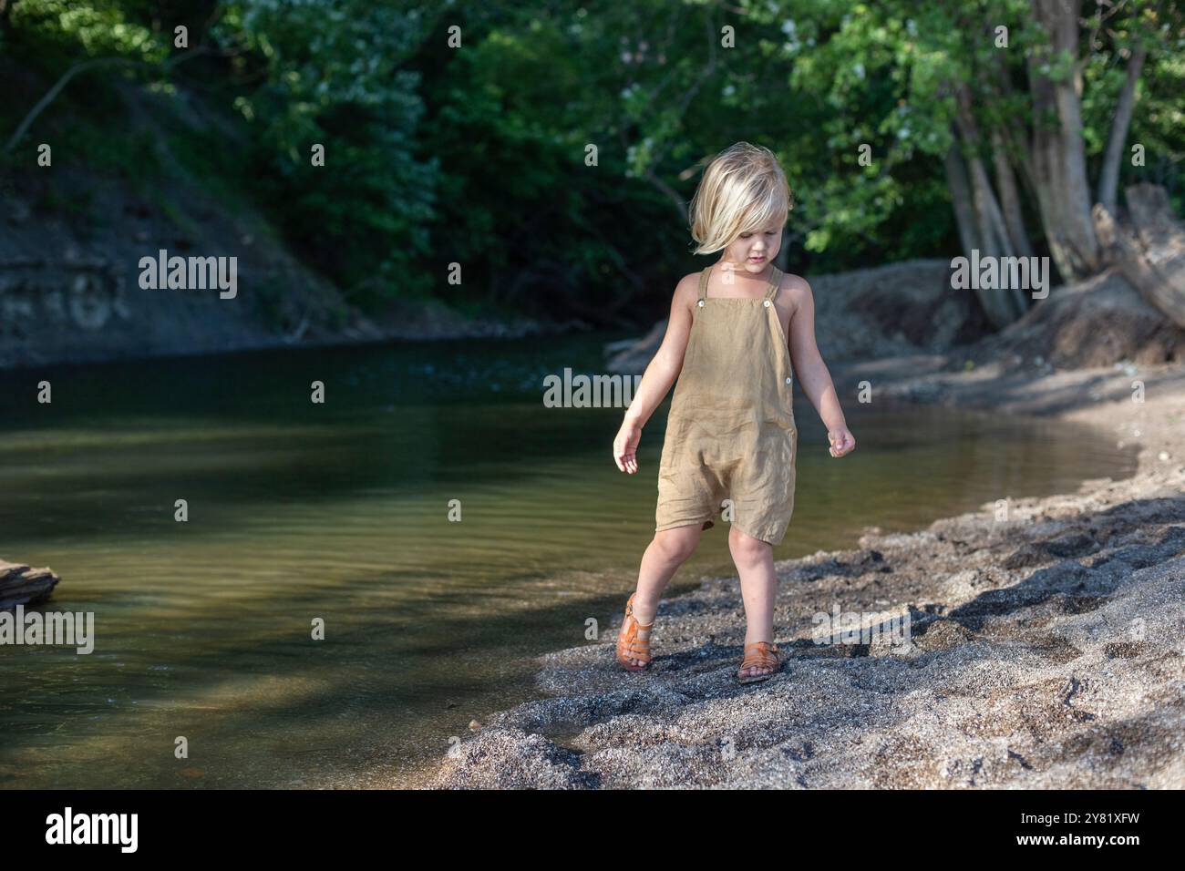 A young child in overalls walks barefoot along a sandy riverbank Stock ...
