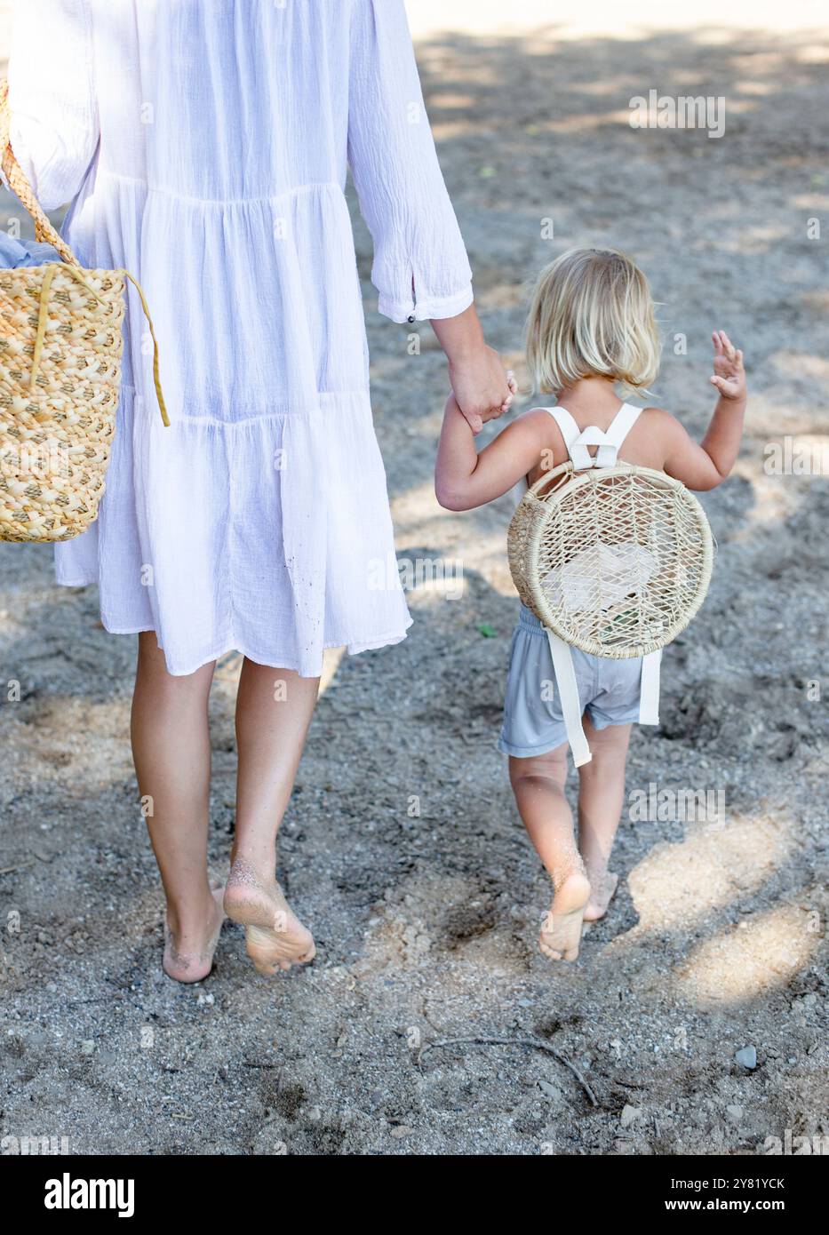 Mother and child walking hand in hand on a sandy path, both carrying ...