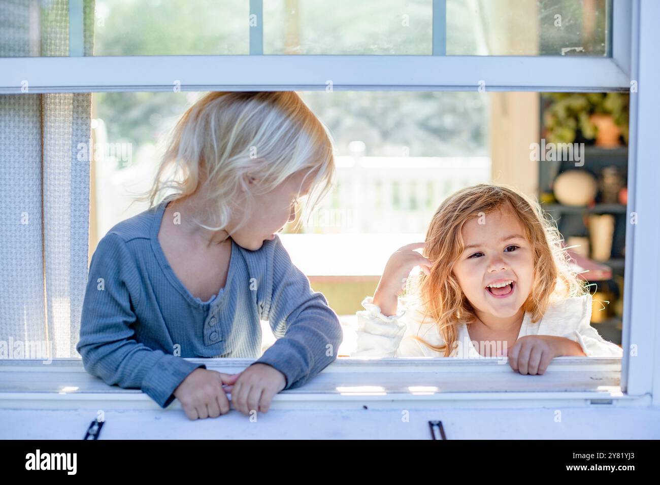 Two young children smile and lean on a windowsill, looking outside on a ...