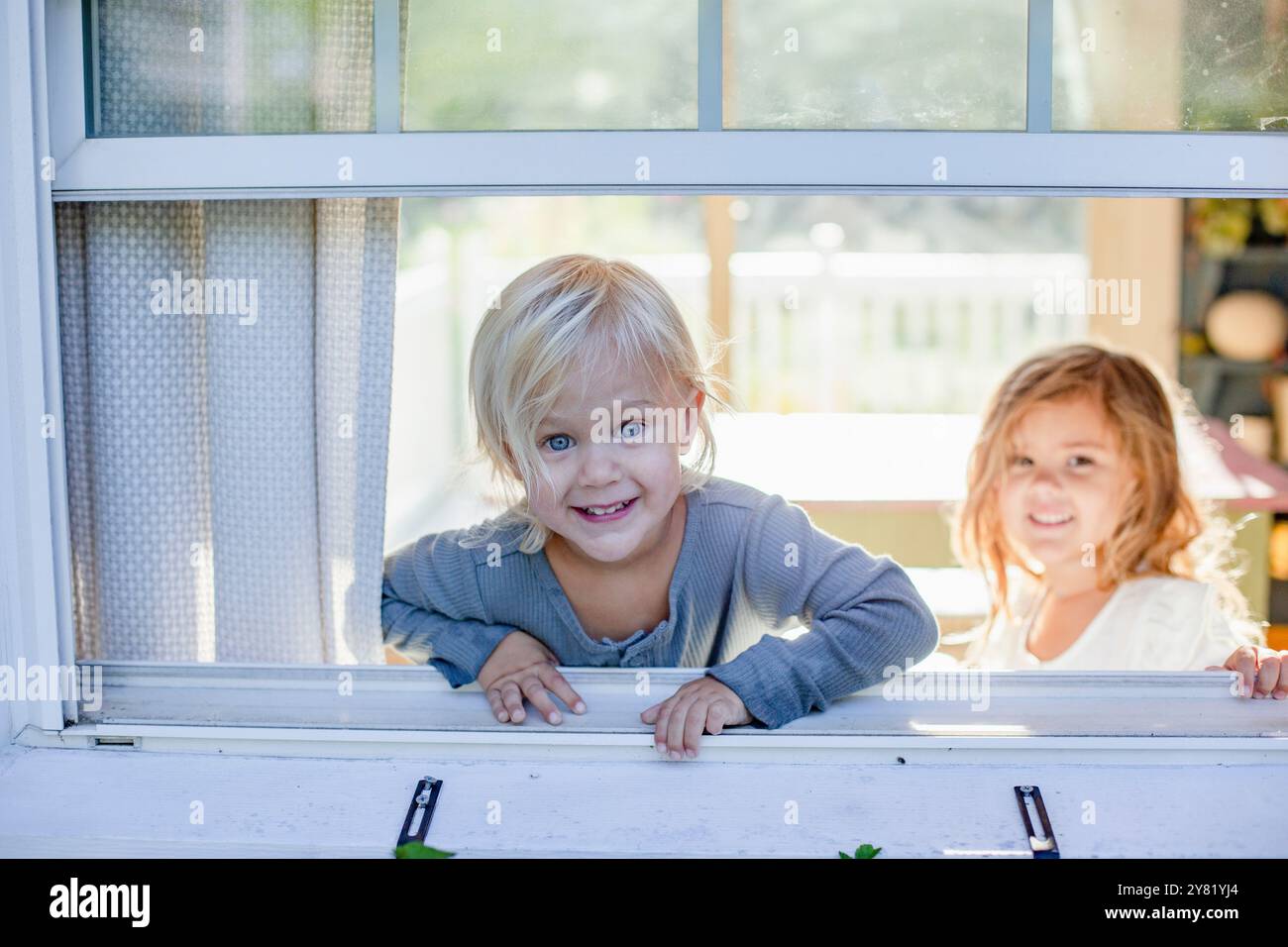 Two smiling children peek out from a white window sill, with sunlight ...