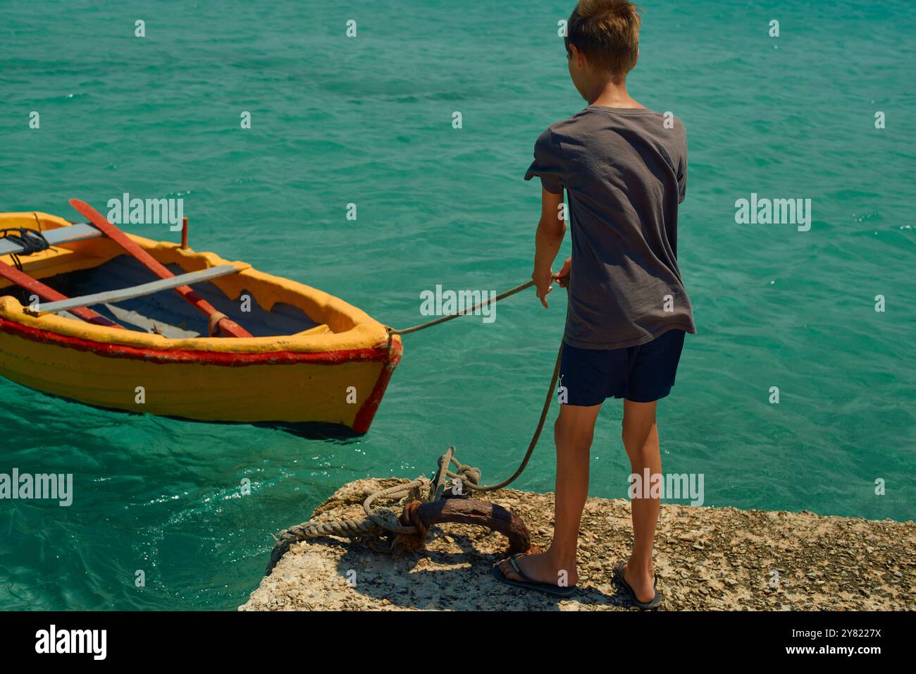 A young boy stands on a jetty pulling a yellow rowboat with turquoise ...