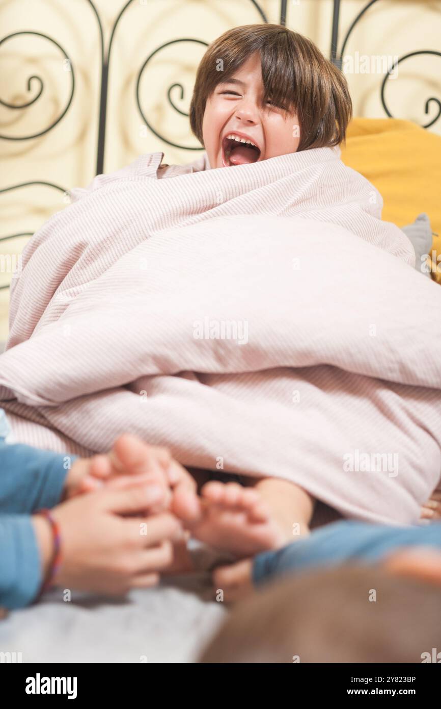 Child covered with a duvet laughing when other children tickle his feet ...