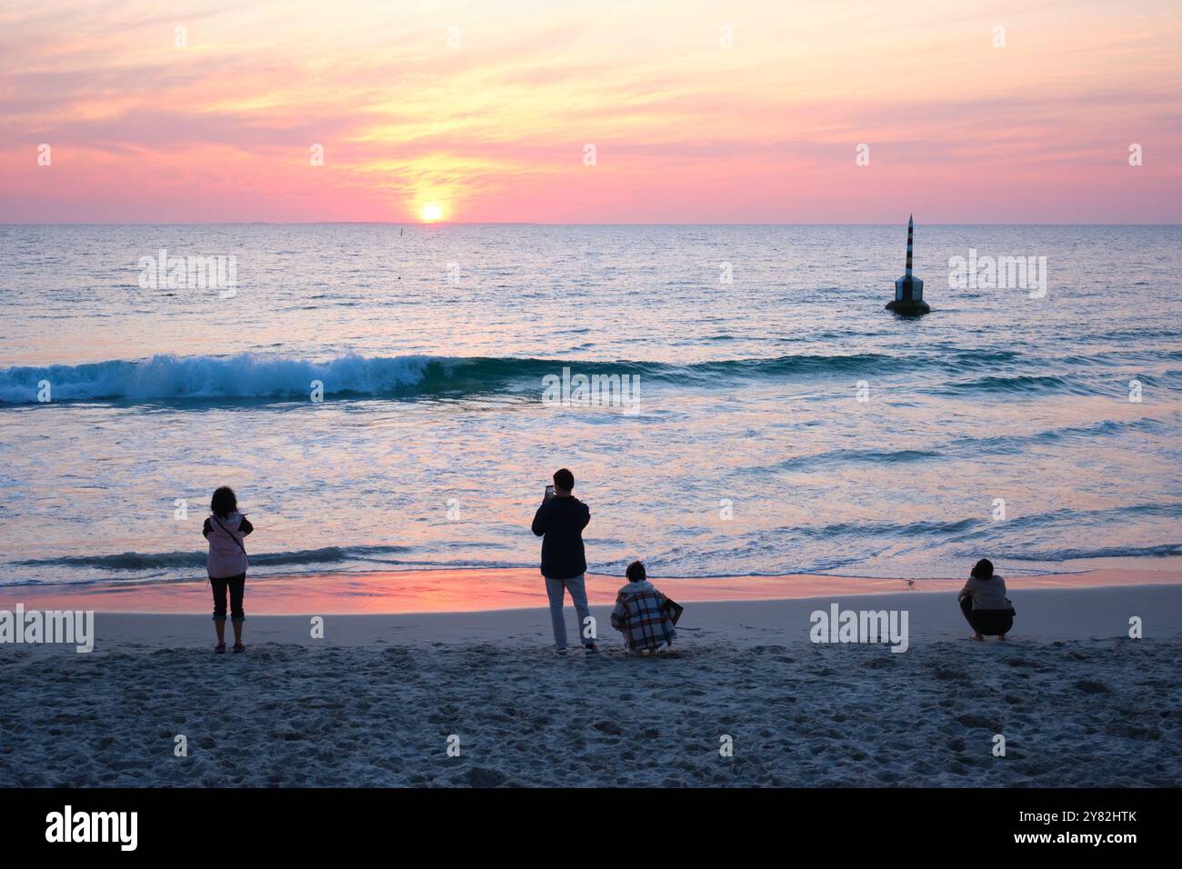 People on the shoreline enjoying photographing the sunset as a wave ...