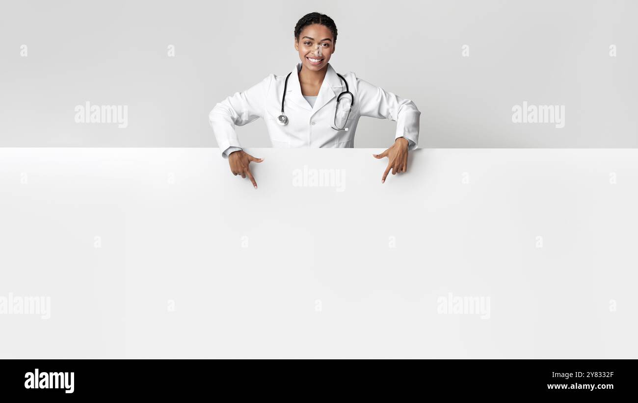 Smiling afro female intern doctor pointing at blank advertisement board ...