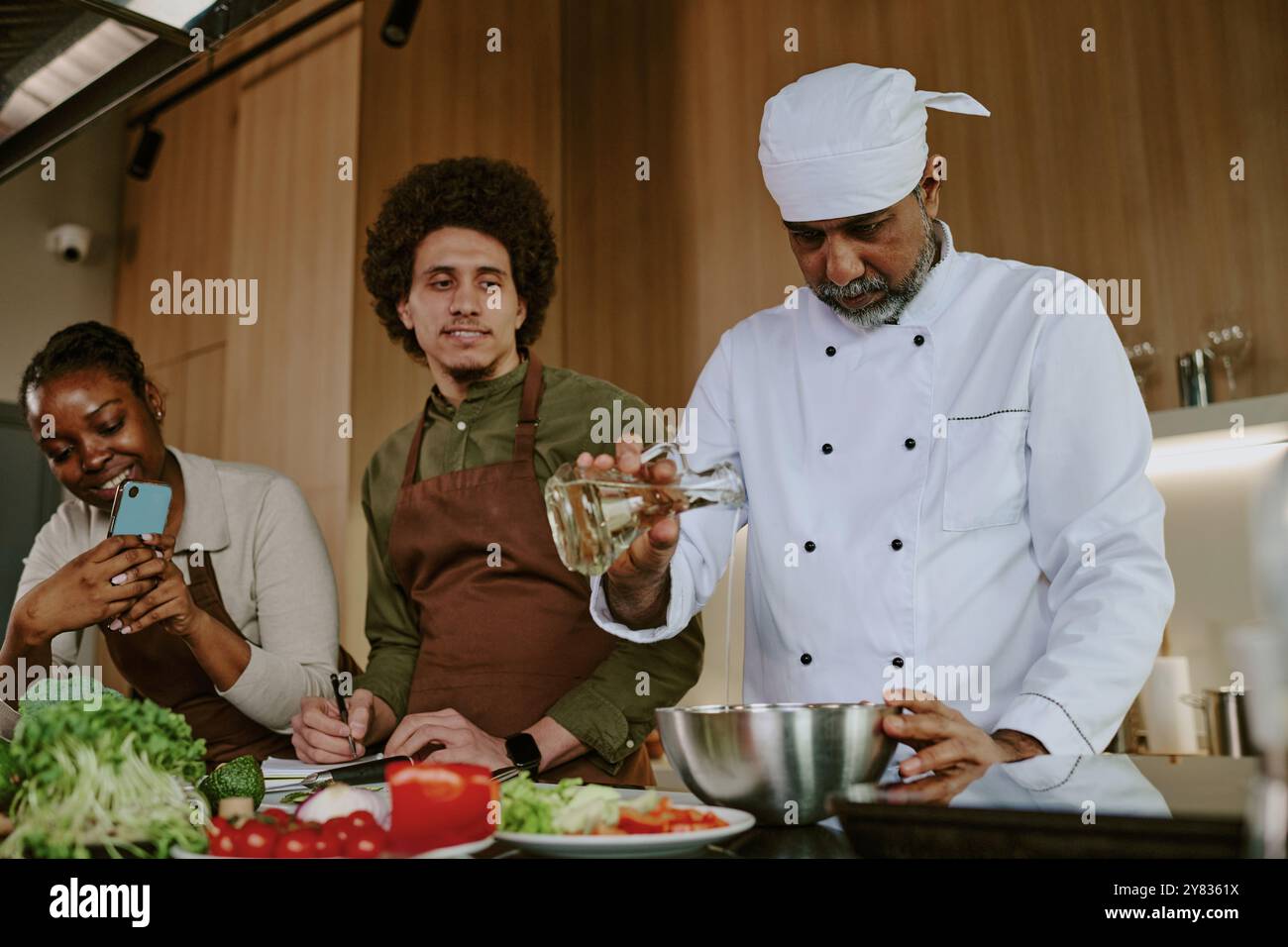 Low angle shot of Indian chef adding vinegar while interns recording ...