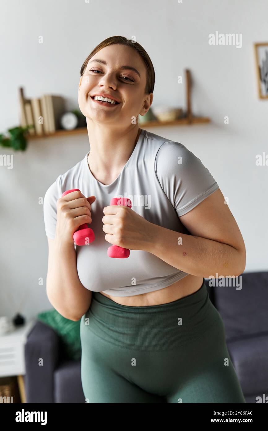 A young sportive woman smiles while exercising at home, lifting pink ...