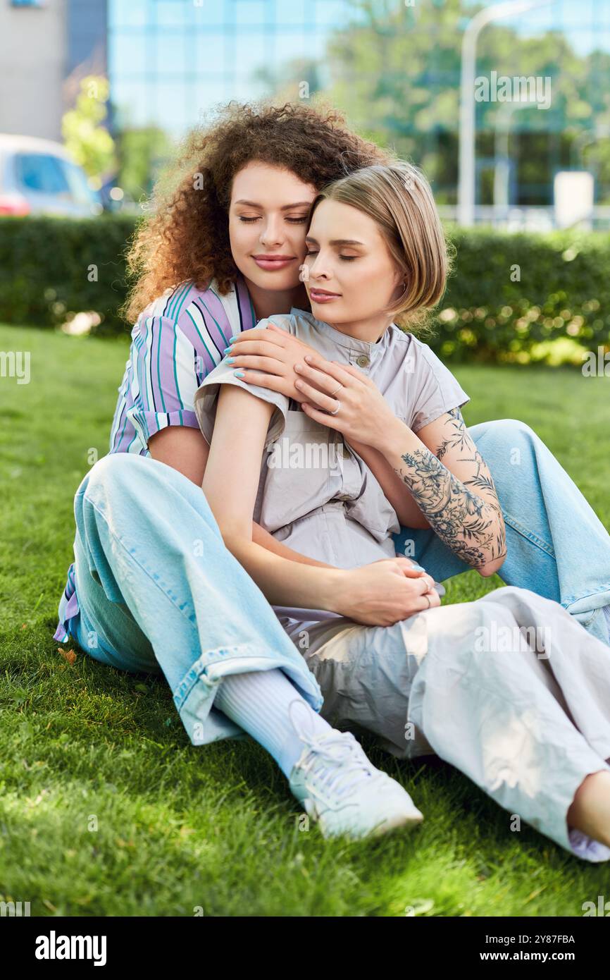 Two young women sit close in a grassy park, sharing moments of joy and ...