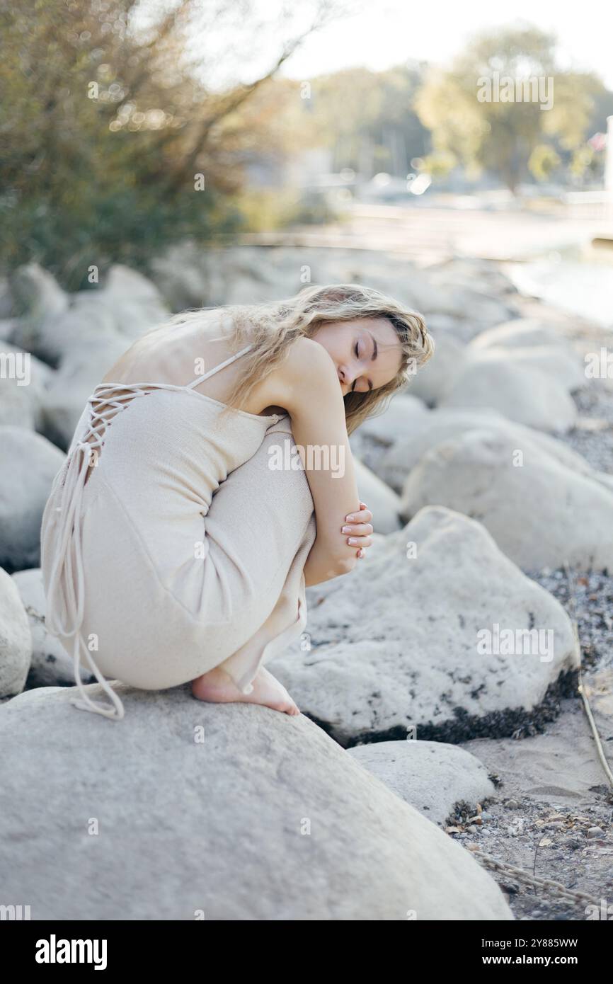 Young lonely sad blonde woman in knitted dress sitting on beach hugging ...