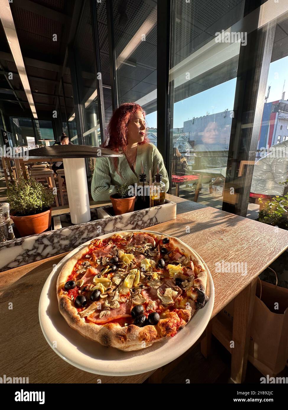 Woman sits by a sunlit window in a cozy restaurant, gazing thoughtfully ...