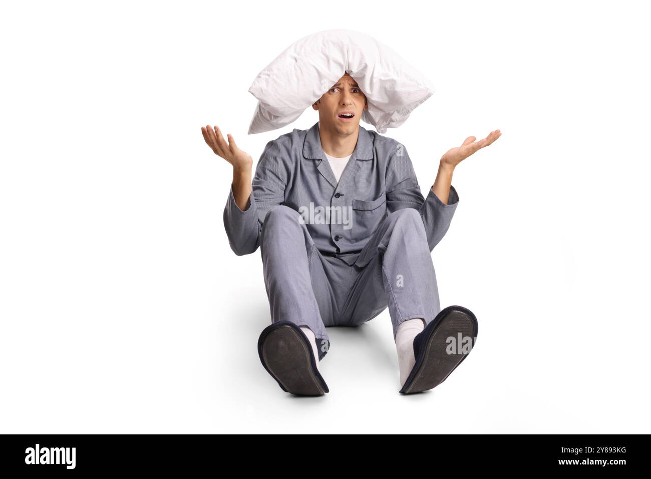 Confused young man in pajamas sitting on the ground with pillow on his ...