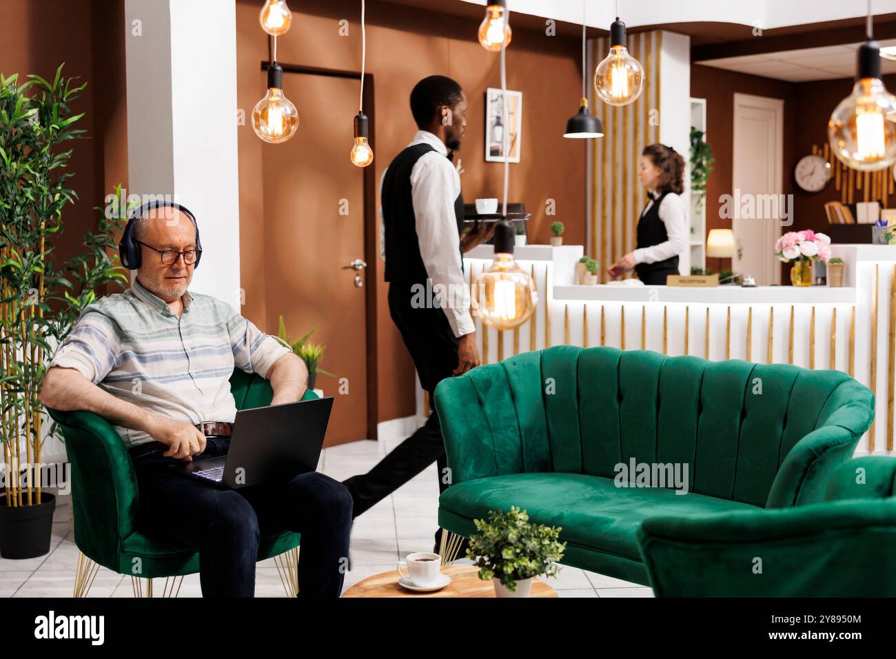 Elderly retired man wears headphones and sits in hotel lobby using his personal computer. Senior ...