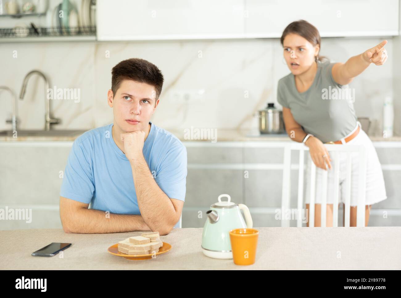 Aggrieved guy sitting at kitchen table while dissatisfied girlfriend ...