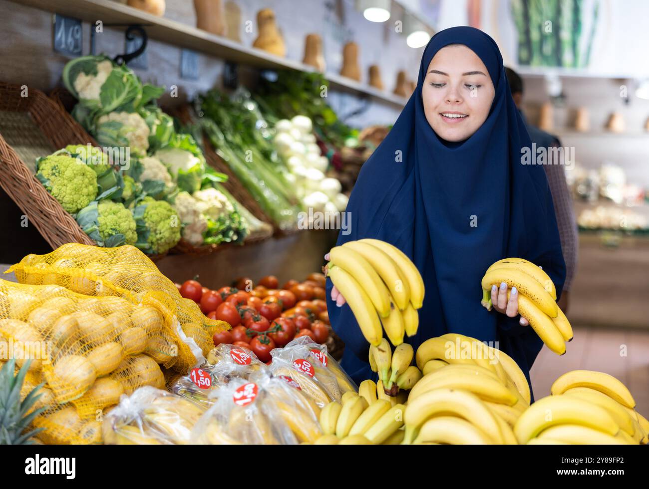Pleased young Muslim woman purchaser choosing bananas in grocery store ...