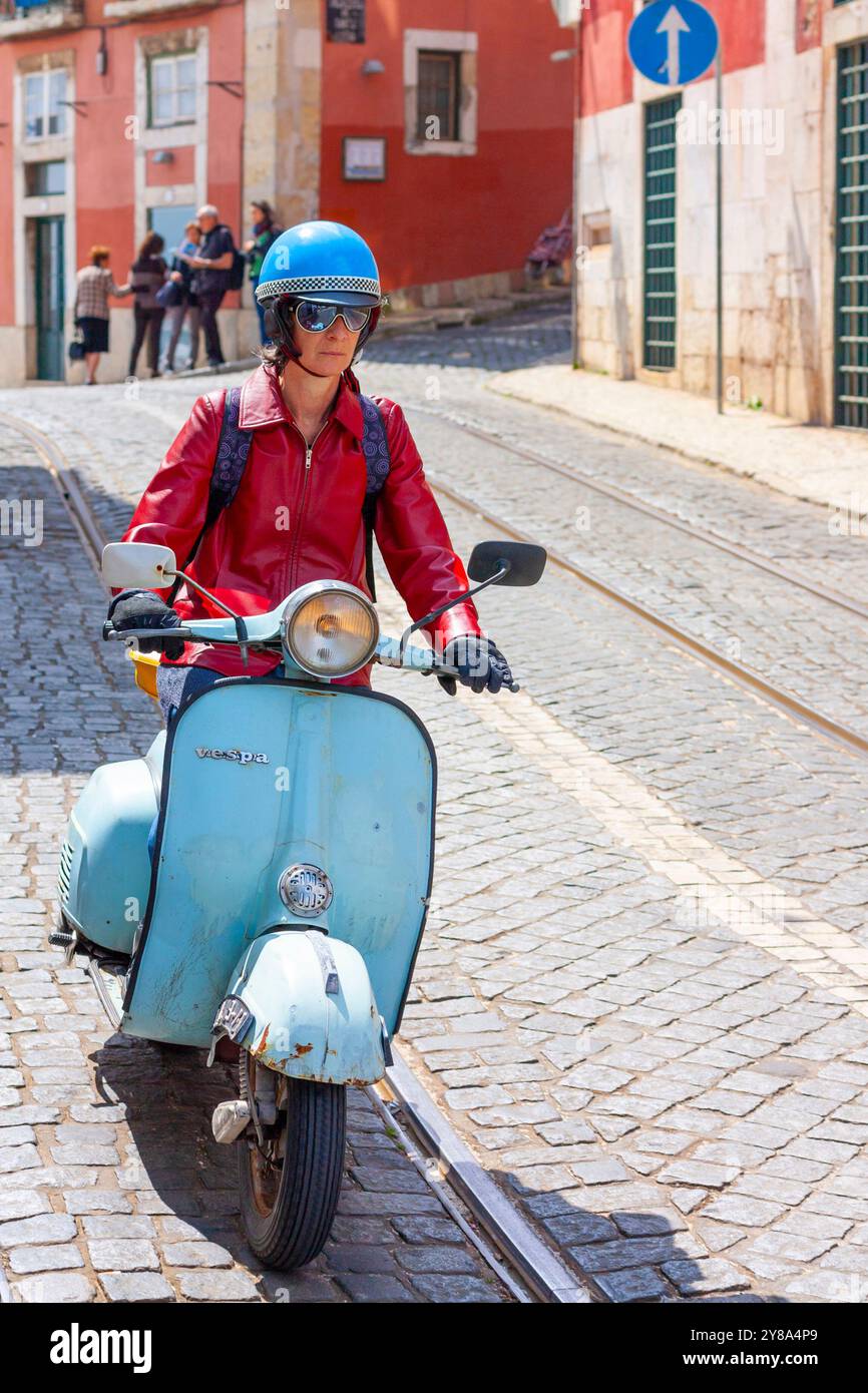 woman in red leather jacket riding a vintage pale blue Piaggio Vespa scooter in a cobbled street ...