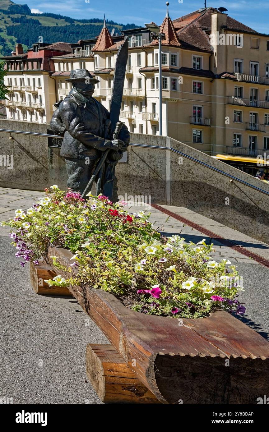 statue, man carrying skis, log planter, flowers, buildings beyond, Alps ...