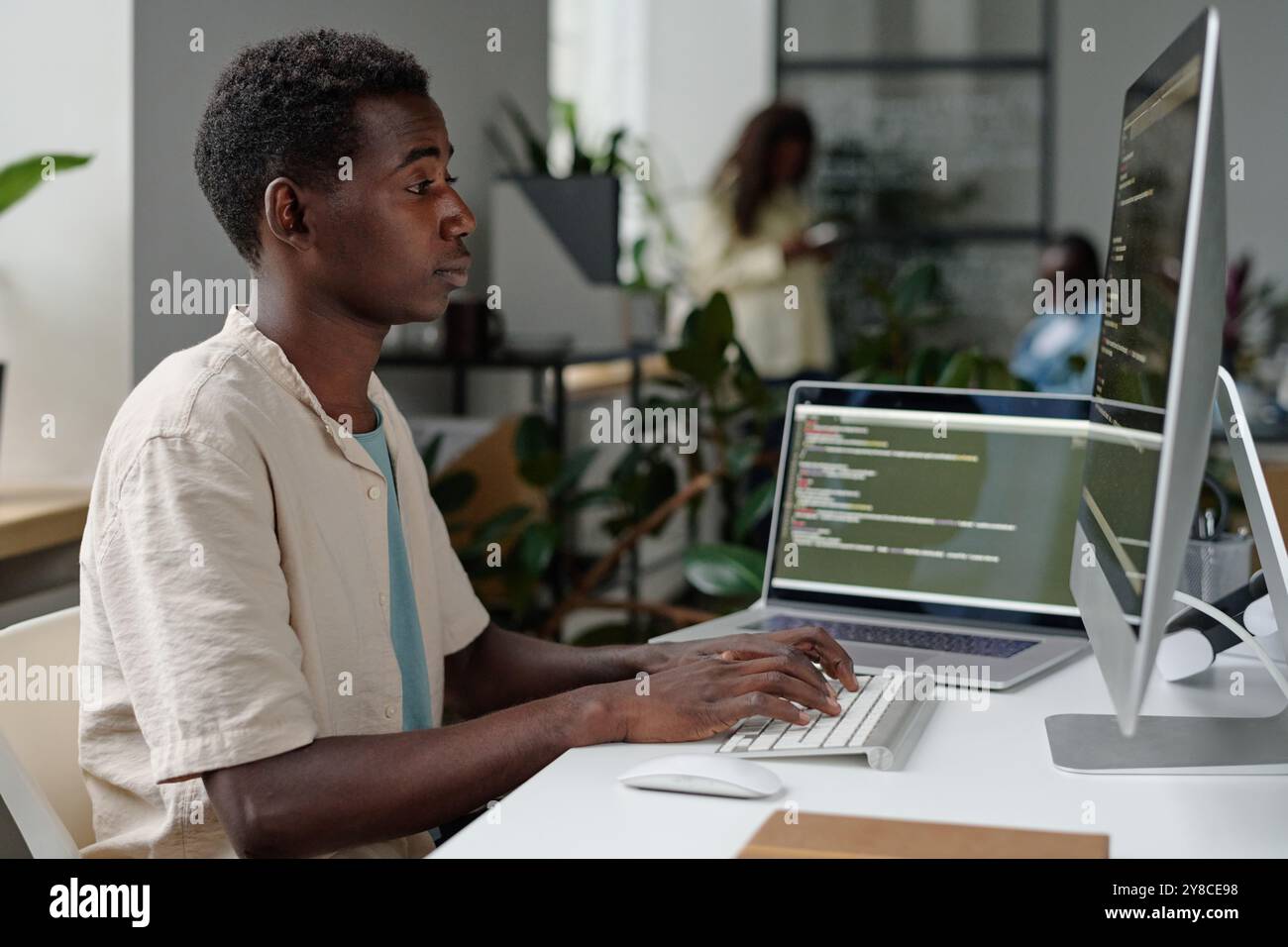 Tired African American programmer coding in cozy office full of plants and electronic devices ...