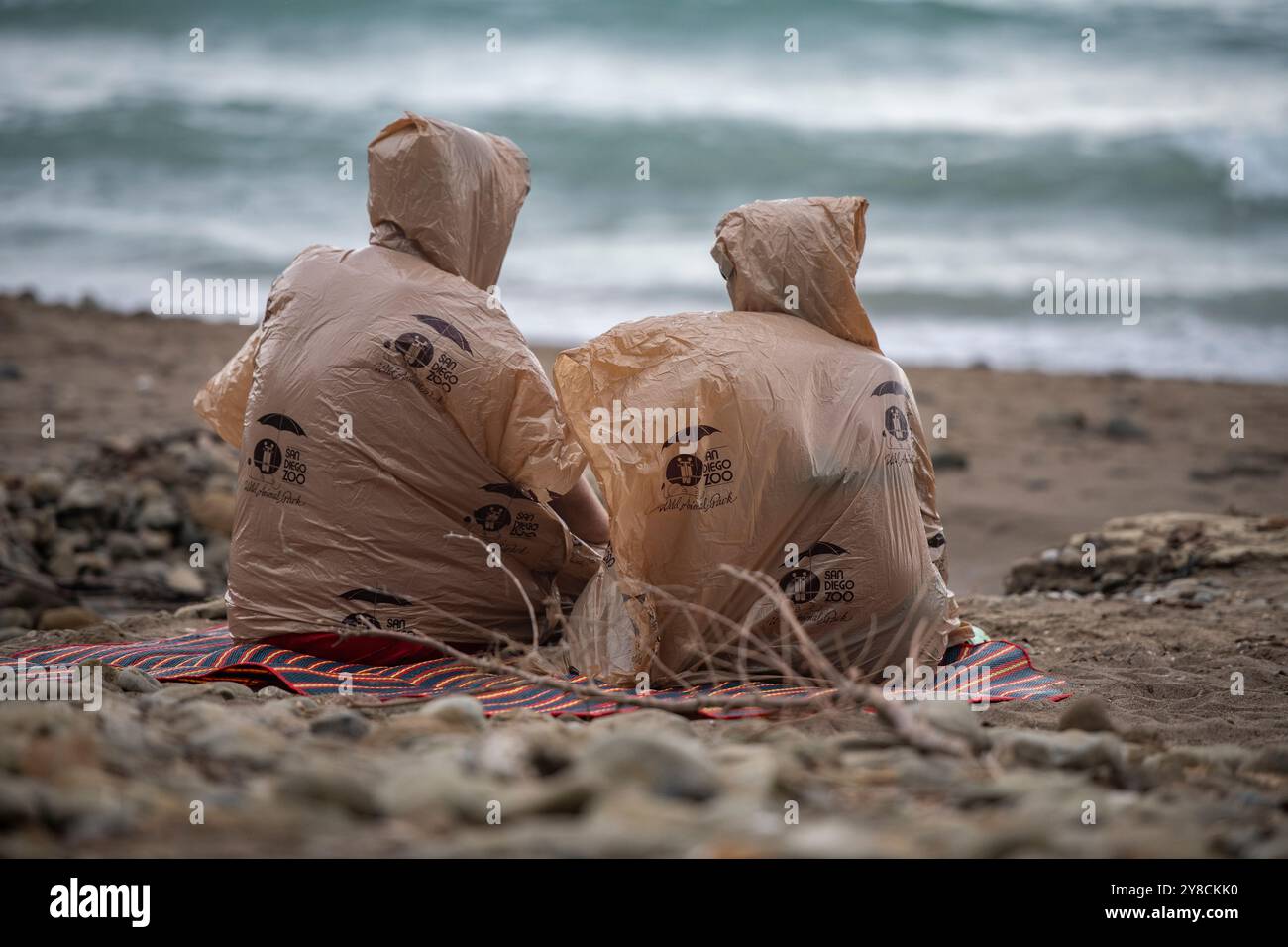 older couple sitting on windswept beach on rainy day wearing ...