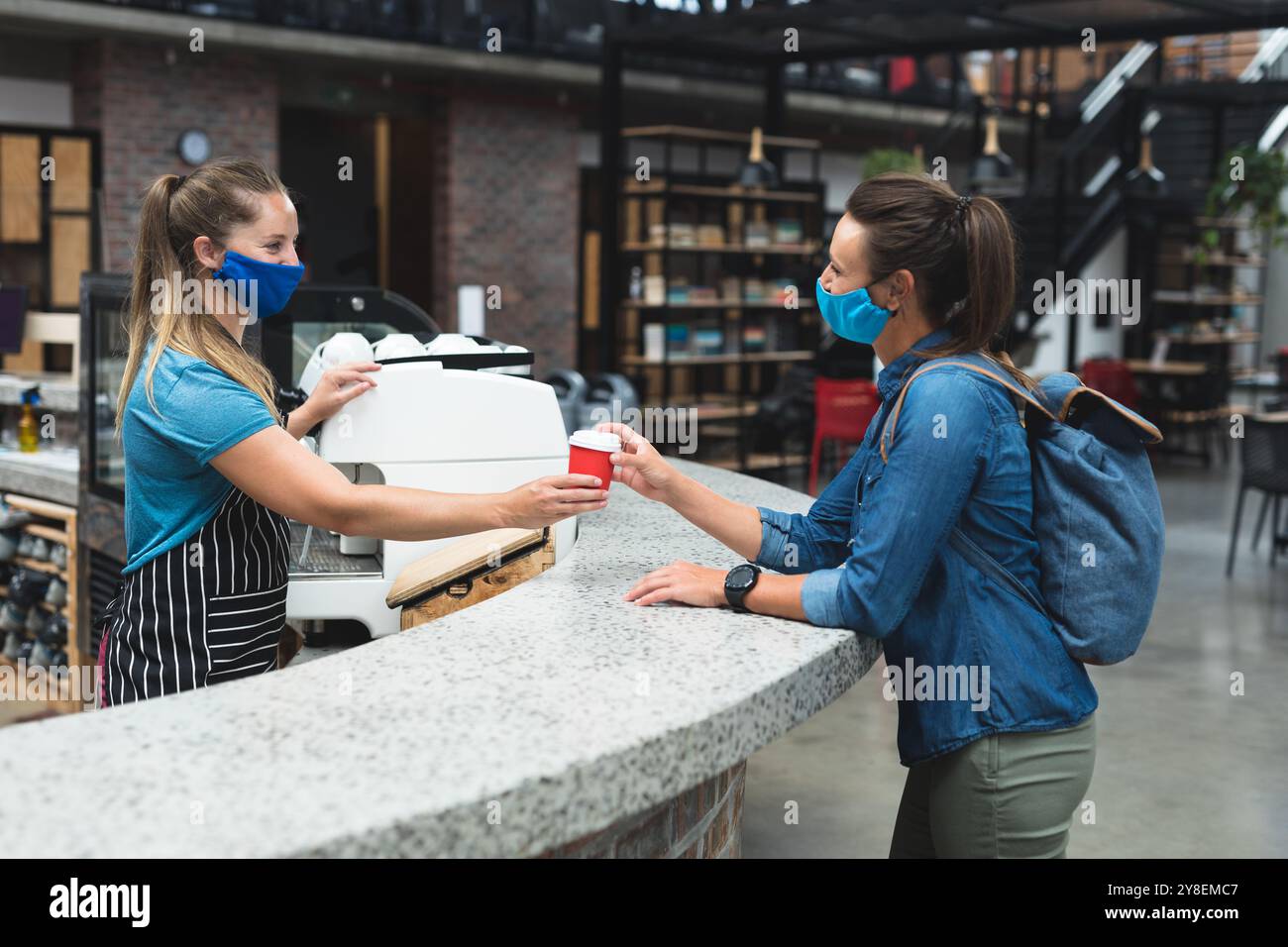 Two happy caucasian women wearing masks passing cup of coffee over ...