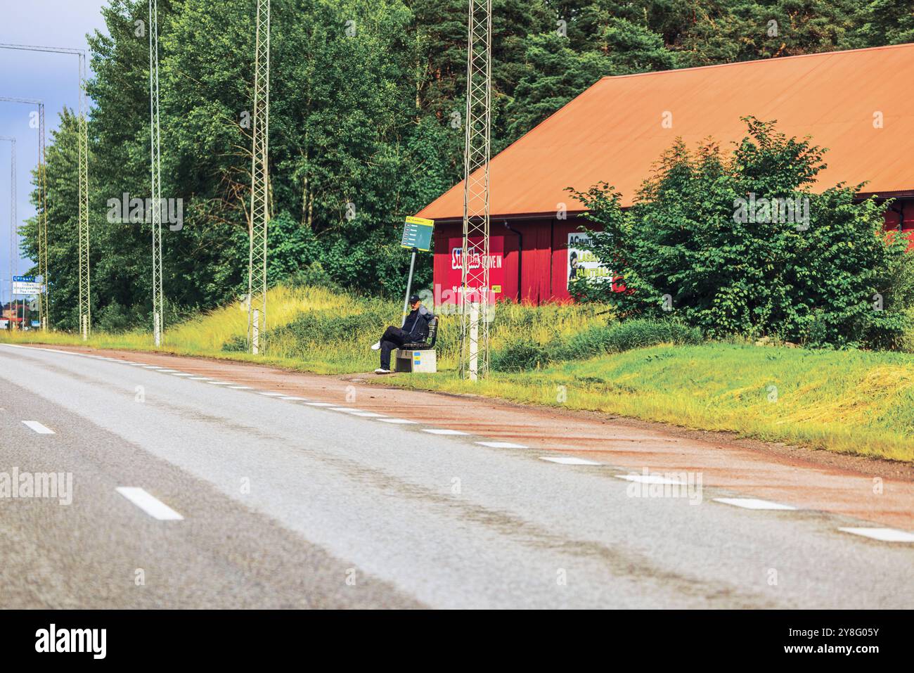 Person sitting alone at rural bus stop near red building, surrounded by ...