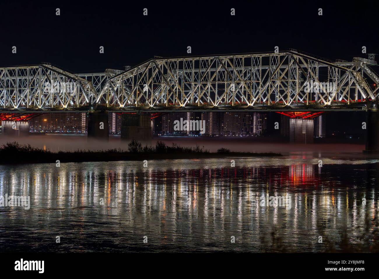 A steel bridge illuminated at night spans across a river, with ...
