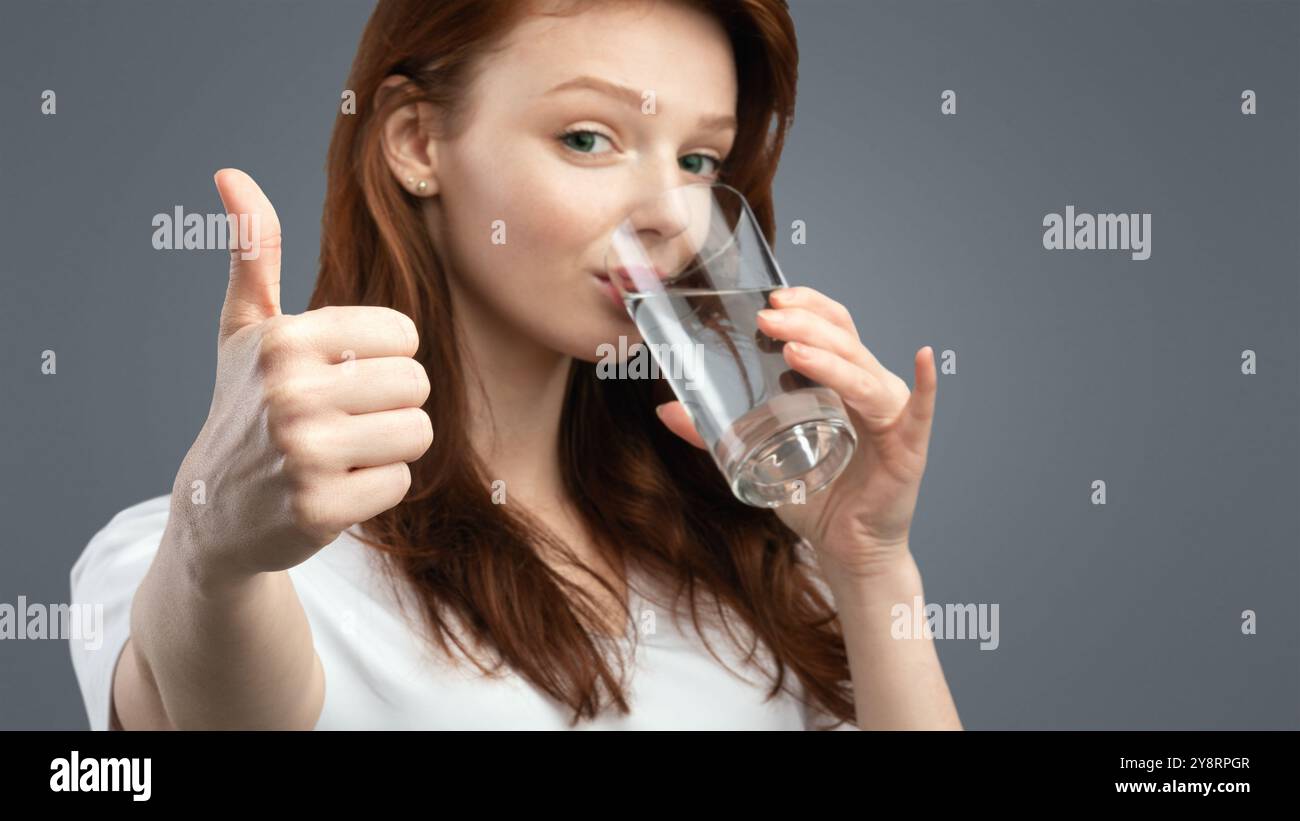 Ginger-Haired Young Lady Drinking Water Gesturing Thumbs-Up Over Gray ...
