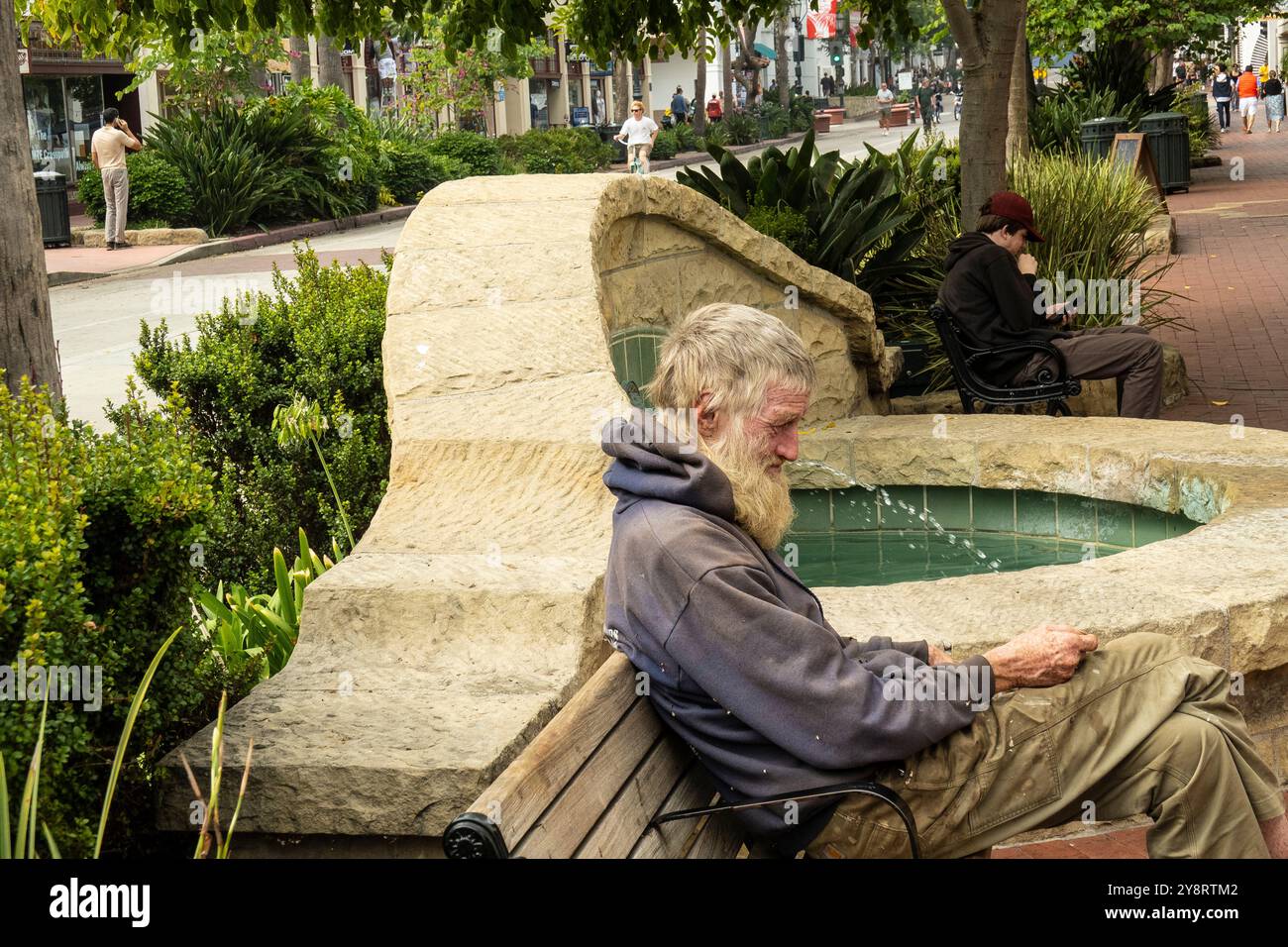 A senior homeless man sits in a chair by a fountain with a stream of ...