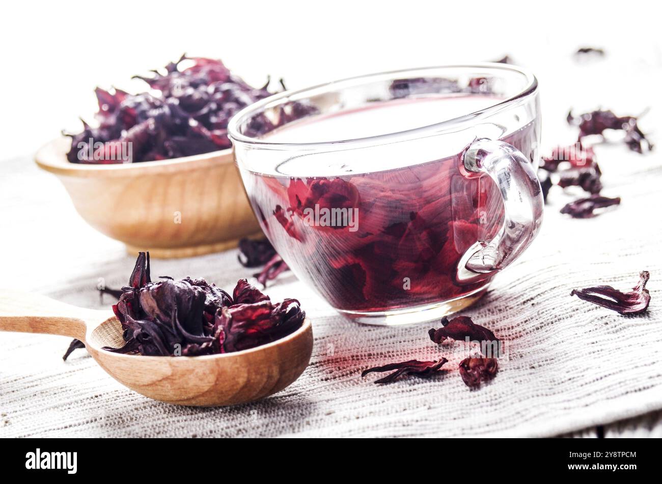 Closeup view at wooden bowl tea cup and spoon of dry hibiscus petals on linen cloth background ...