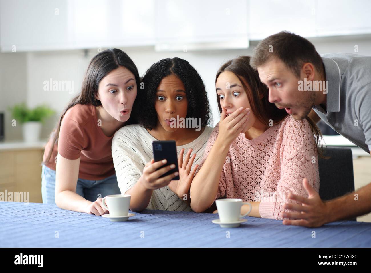 Four shocked interracial friends checking phone in the kitchen at home ...