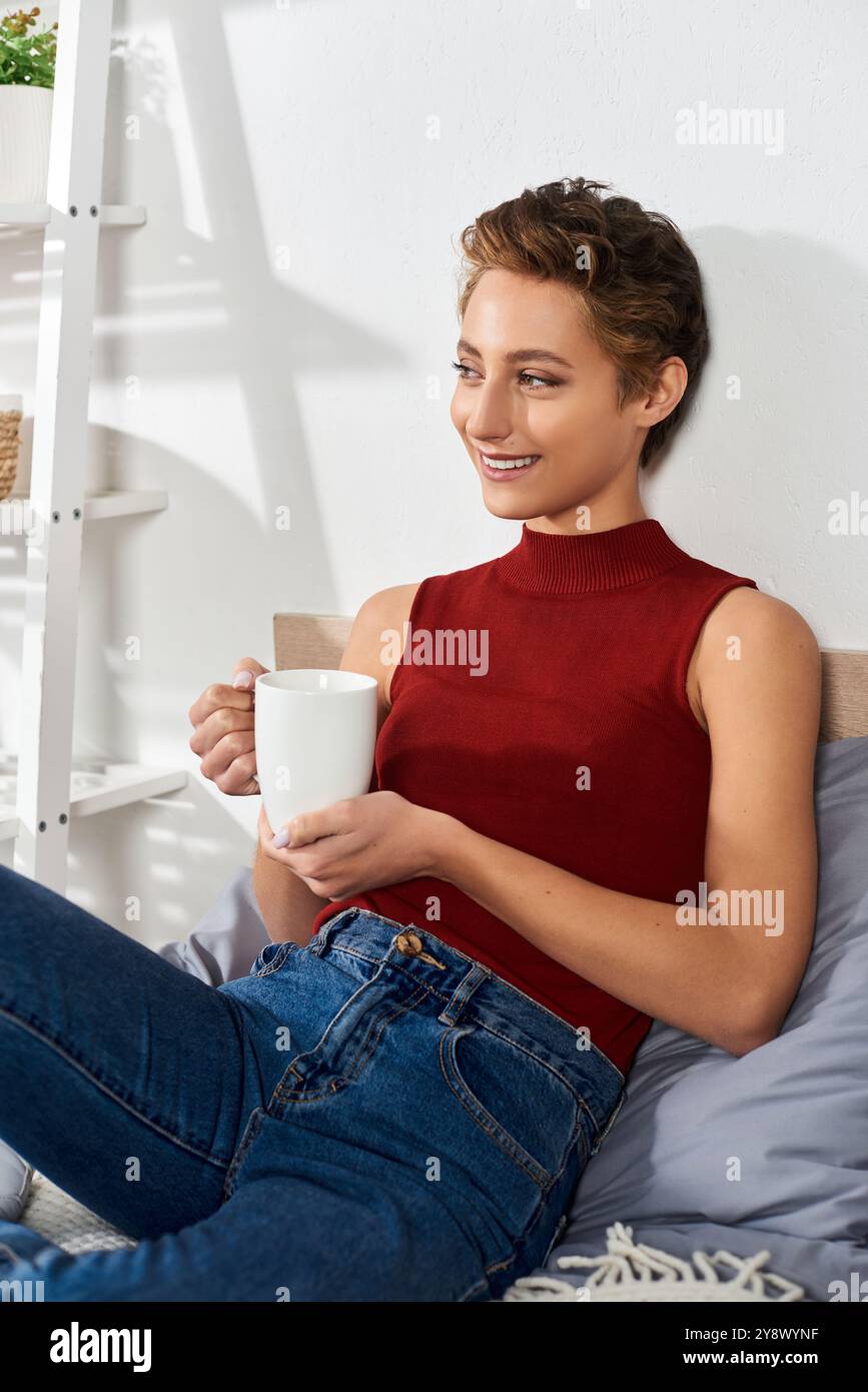 A young woman in a red tank top sips coffee while lounging at home ...